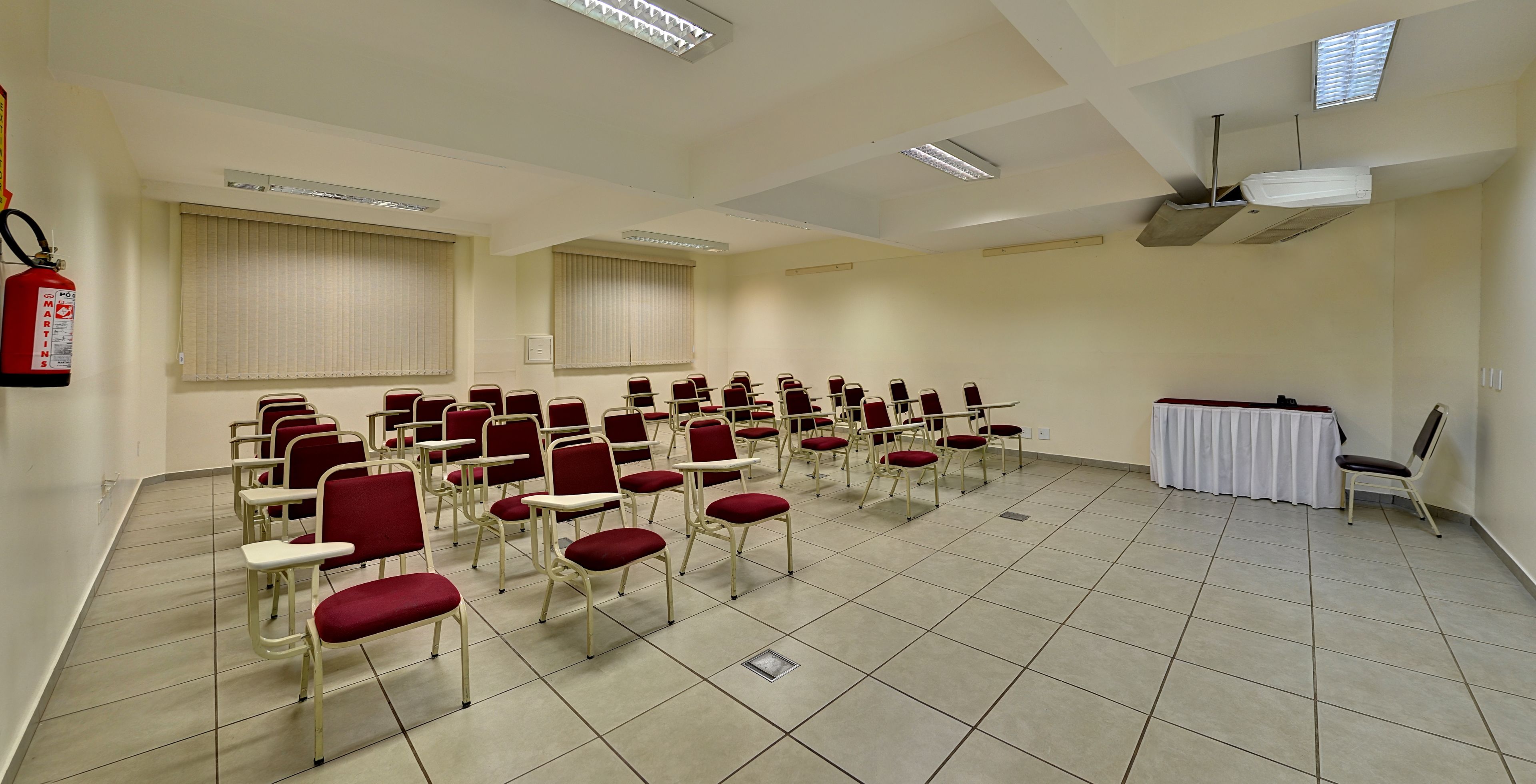 Uma sala de conferências com fileiras de cadeiras vermelhas e mesas, um extintor de incêndio e ventiladores no teto.
