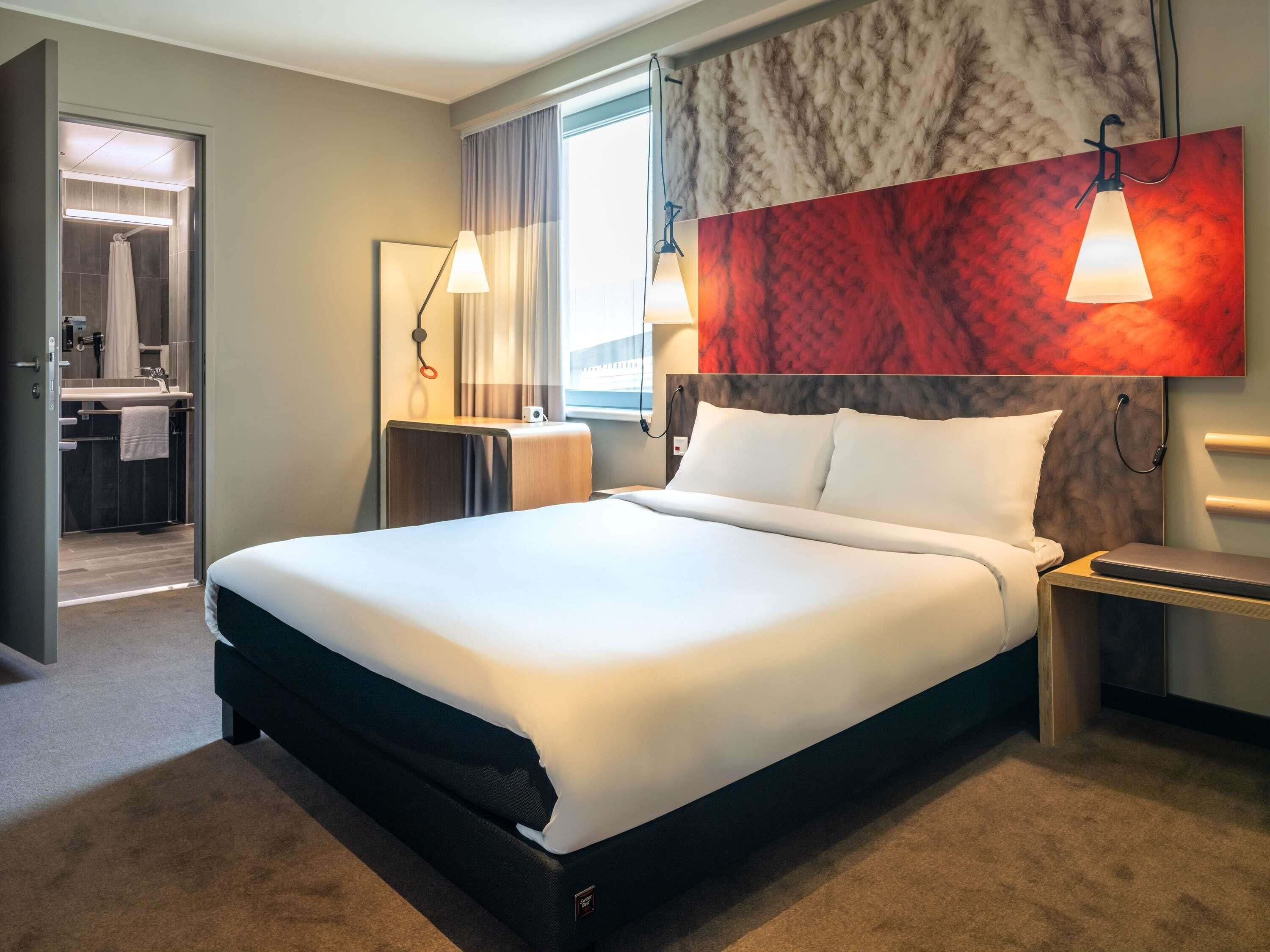 Quarto de hotel com uma cama grande, uma escrivaninha com um laptop, uma mesa de cabeceira, uma janela com cortinas, um banheiro com chuveiro e pia, e uma cabeceira com um desenho estampado em vermelho e cinza.