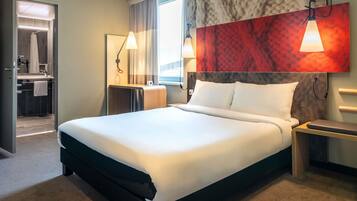Quarto de hotel com uma cama grande, uma escrivaninha com um laptop, uma mesa de cabeceira, uma janela com cortinas, um banheiro com chuveiro e pia, e uma cabeceira com um desenho estampado em vermelho e cinza.
