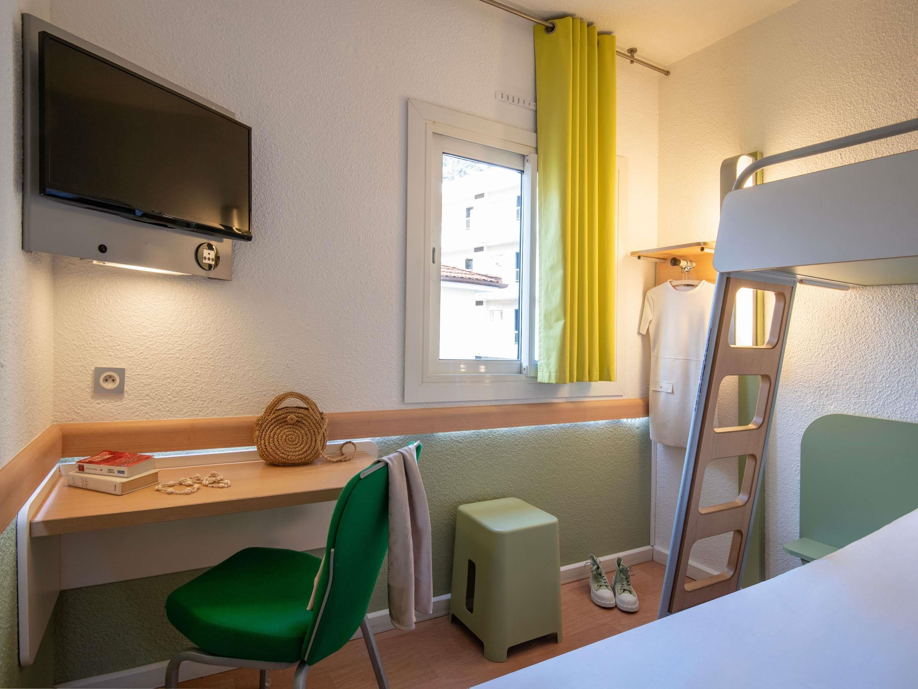 Quarto de hotel com uma cama, escrivaninha com cadeira, televisão na parede, janela com cortinas e uma cama beliche.