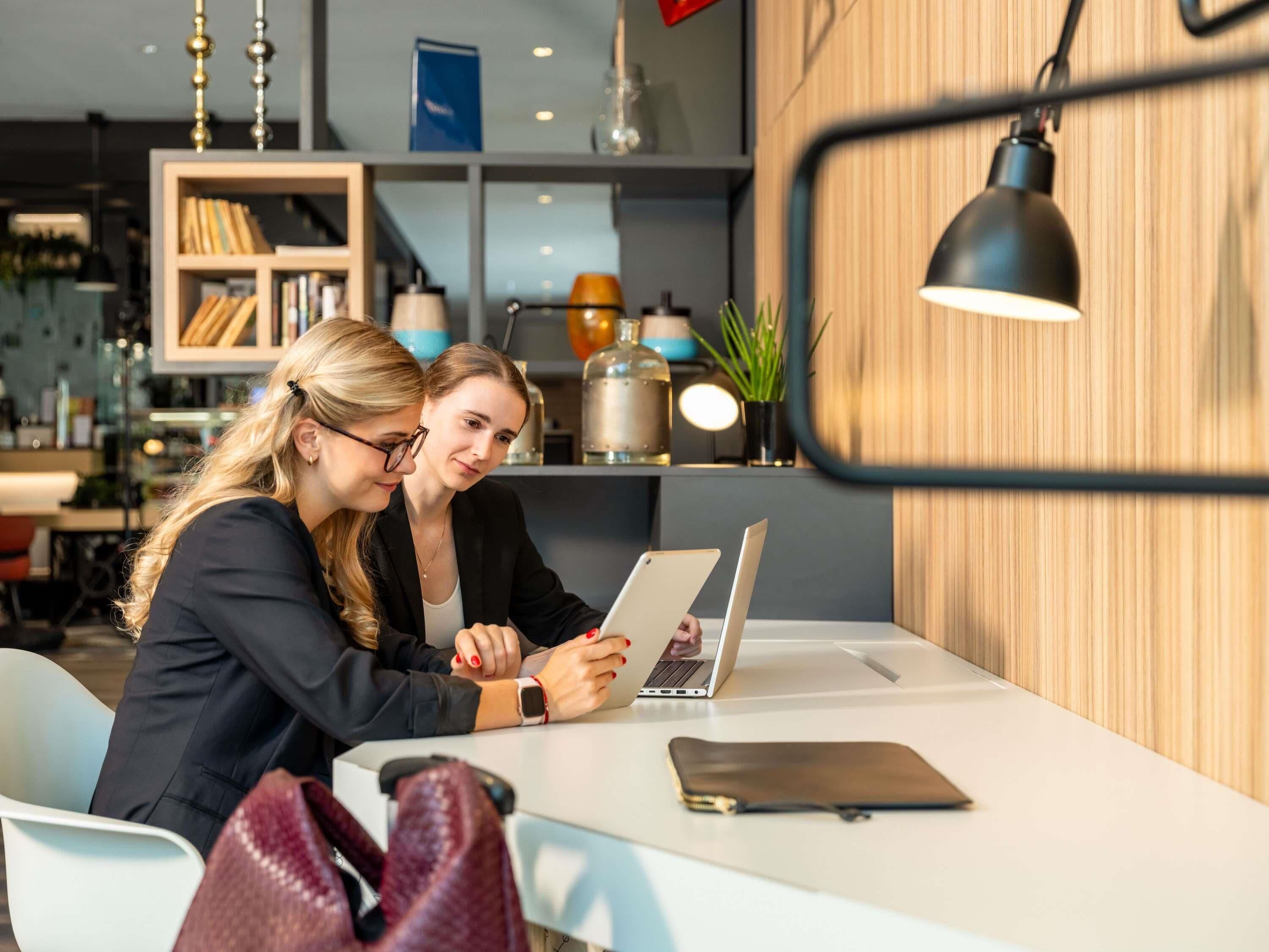 Duas pessoas à mesa com um laptop e um tablet, uma luminária e uma estante com livros e objetos decorativos ao fundo.