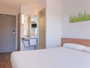 Quarto de hotel com uma cama, banheiro com pia e espelho, e uma obra de arte na parede.