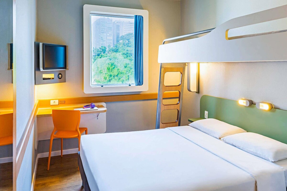 Quarto de hotel com uma cama de solteiro, uma escrivaninha com cadeira, televisão e janela com vista para edifícios e árvores.