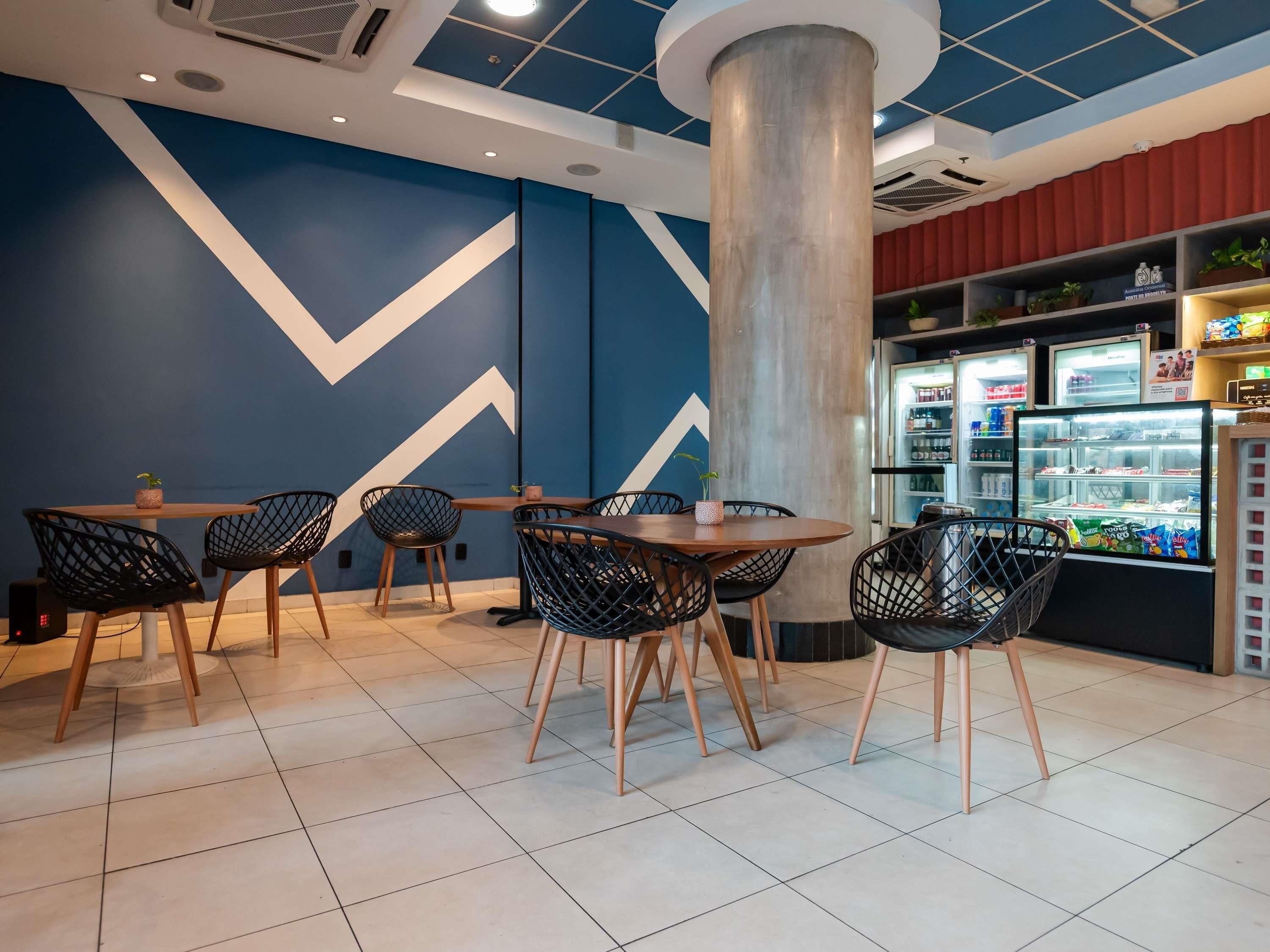 Um café com decoração moderna, destacando um pilar grande, parede azul com padrão ziguezague branco e um refrigerador cheio de bebidas.