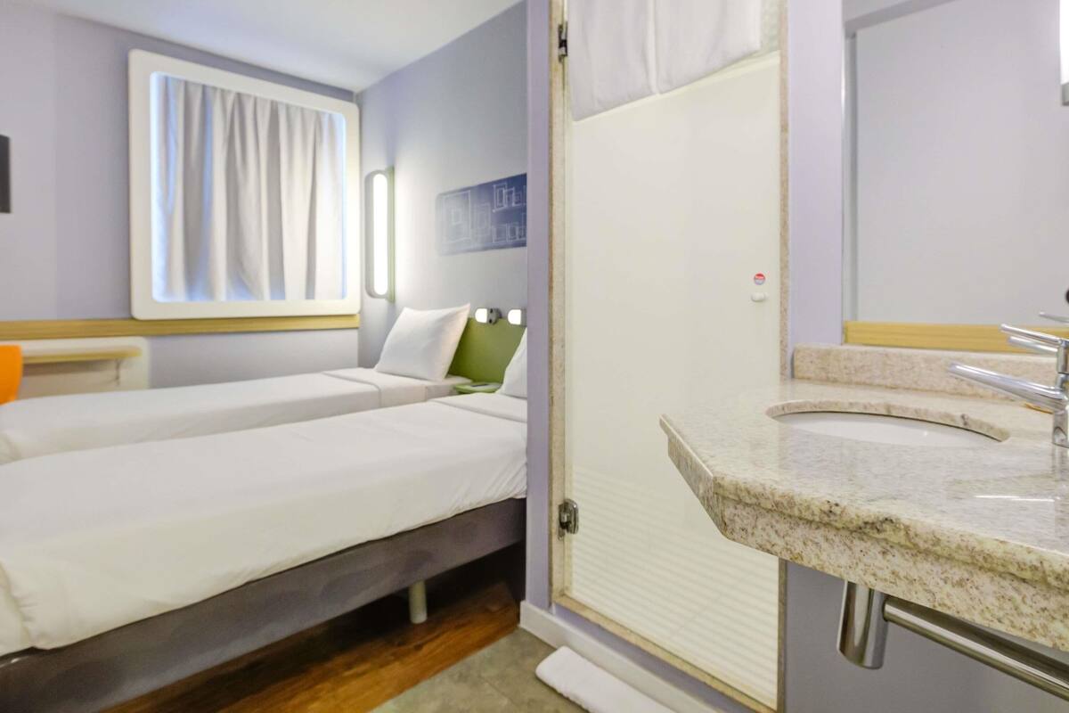 Quarto de hotel com duas camas, banheiro com pia e espelho, e janela com cortinas.