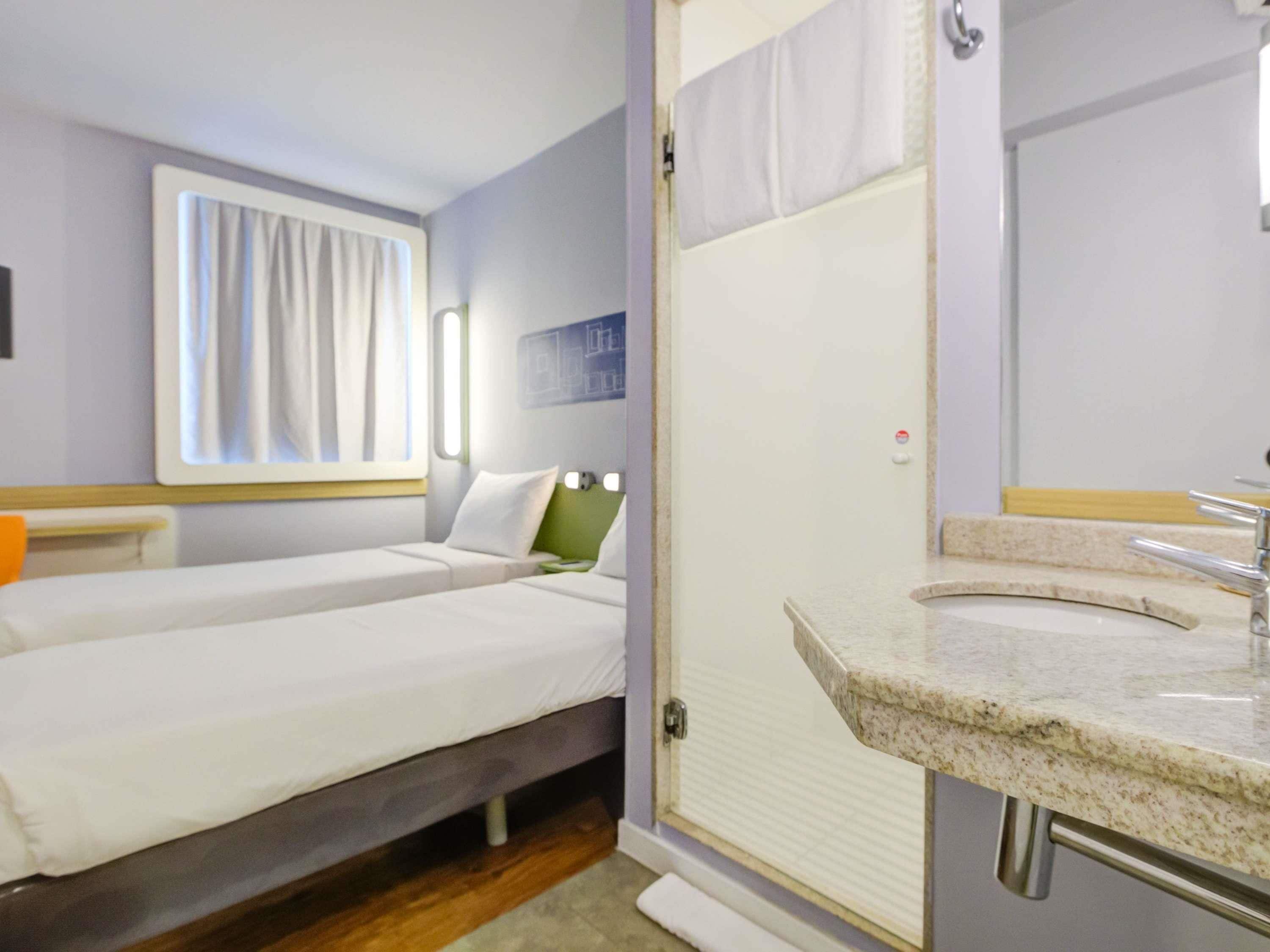 Quarto de hotel com duas camas, banheiro com pia e espelho, e janela com cortinas.