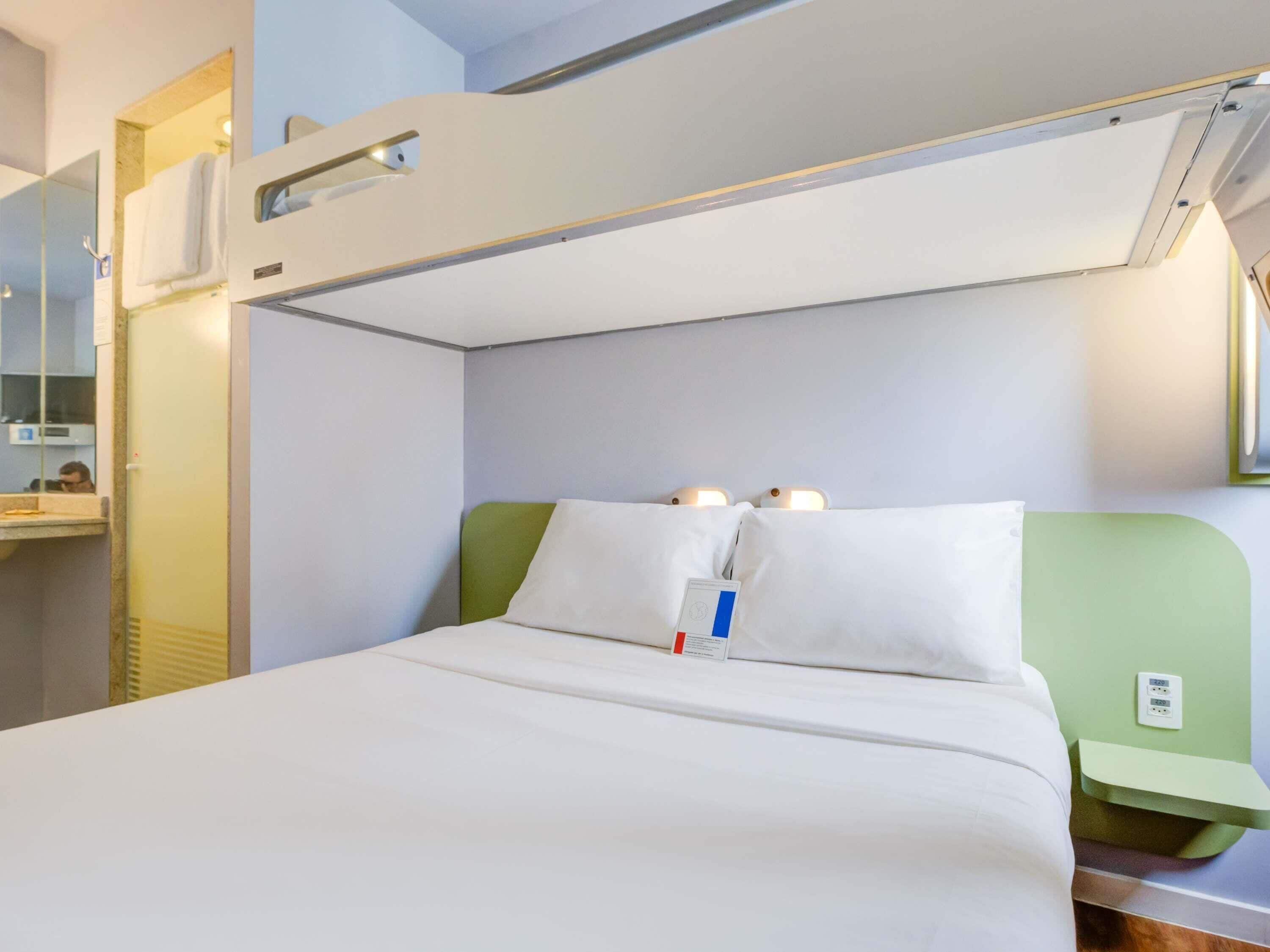 Quarto de hotel com cama de beliche, mesa de cabeceira, espelho e janela.