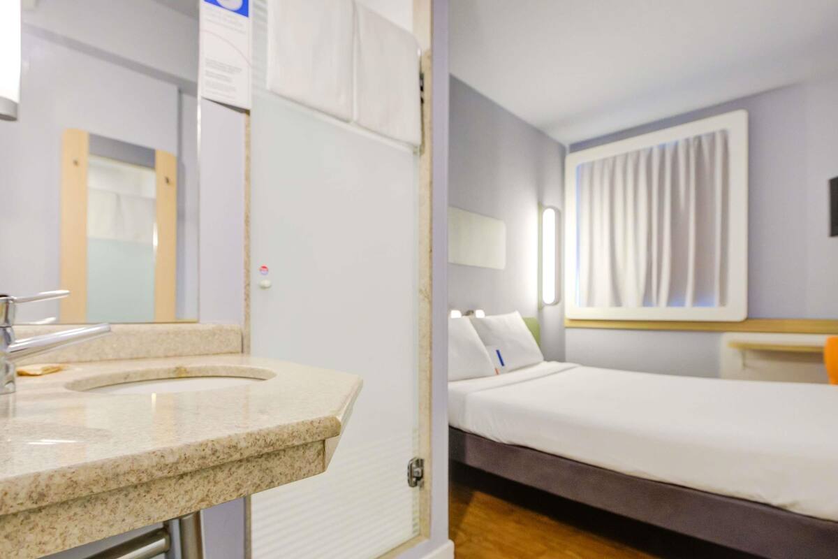 Quarto de hotel com banheiro, cama, mesa de cabeceira e janela com cortinas.