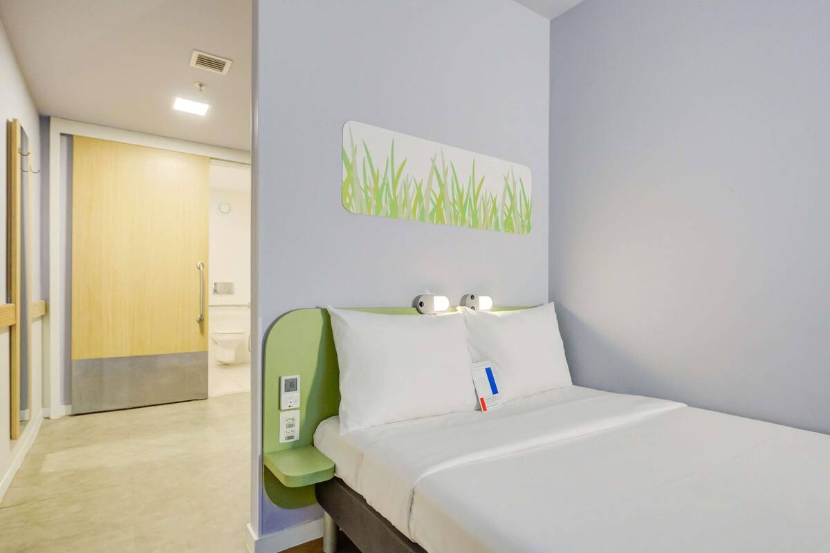 Quarto de hotel com cama, mesa de cabeceira e luminária fixada na parede.