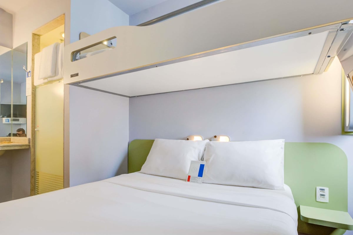 Quarto de hotel com cama de beliche, mesa de cabeceira, espelho e janela.