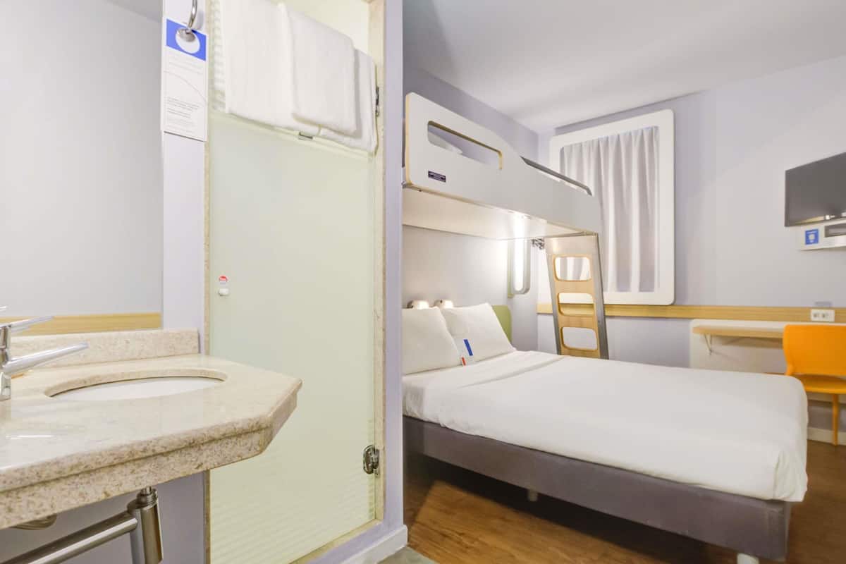Quarto de hotel com cama beliche, banheiro com pia e espelho, escrivaninha e televisão.