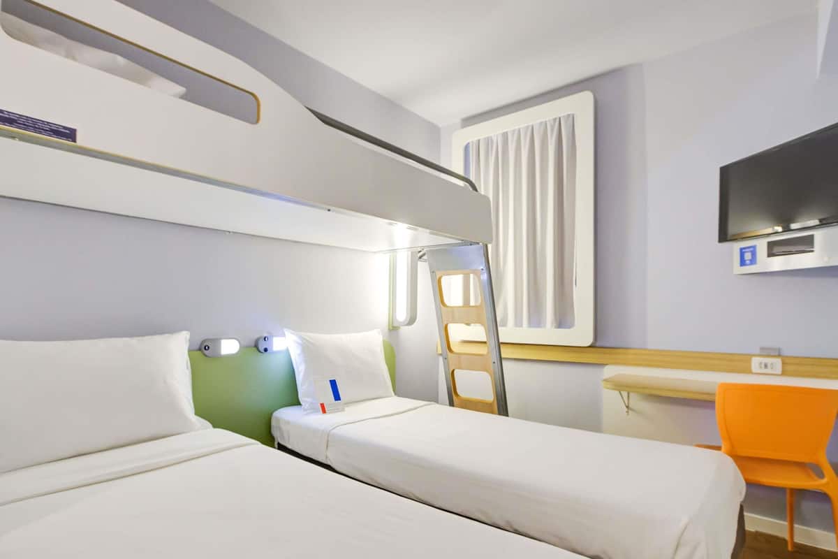 Quarto de hotel com duas camas, uma escrivaninha, uma cadeira, uma televisão e uma janela com cortinas.