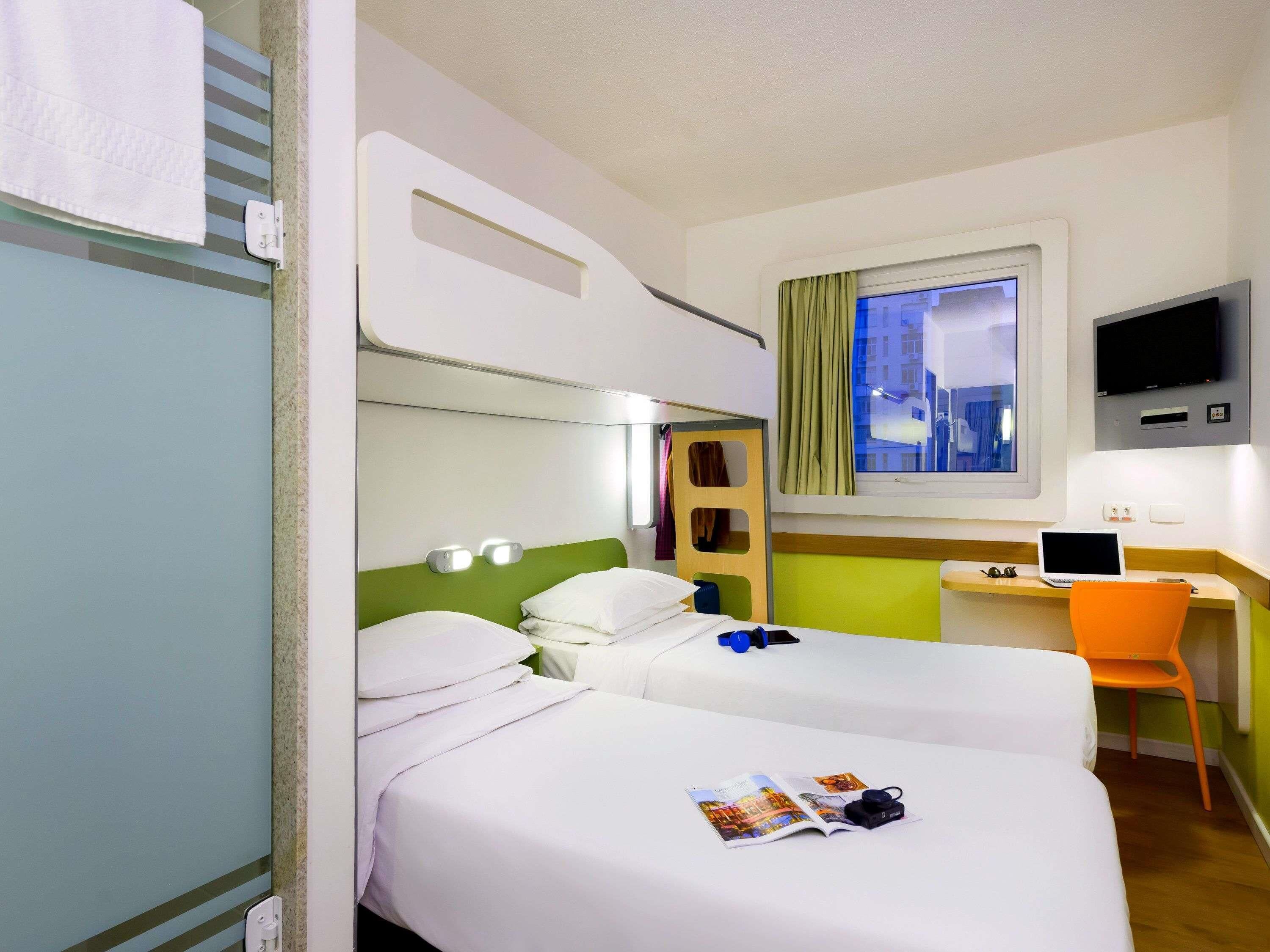 Quarto de hotel com duas camas, uma mesa com um laptop, televisão e janela com vista para edifícios.