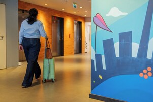 Mulher caminhando com mala em corredor com elevadores e um mural na parede.