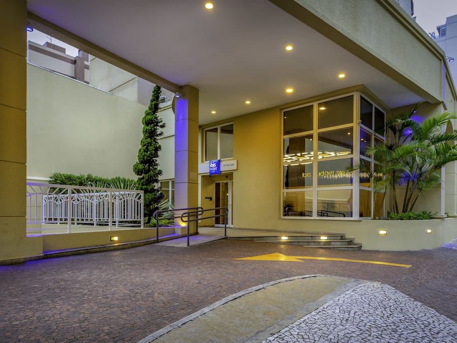 Entrada de hotel com design moderno, incluindo fachada de vidro, escadaria que leva à entrada e caminho bem iluminado.