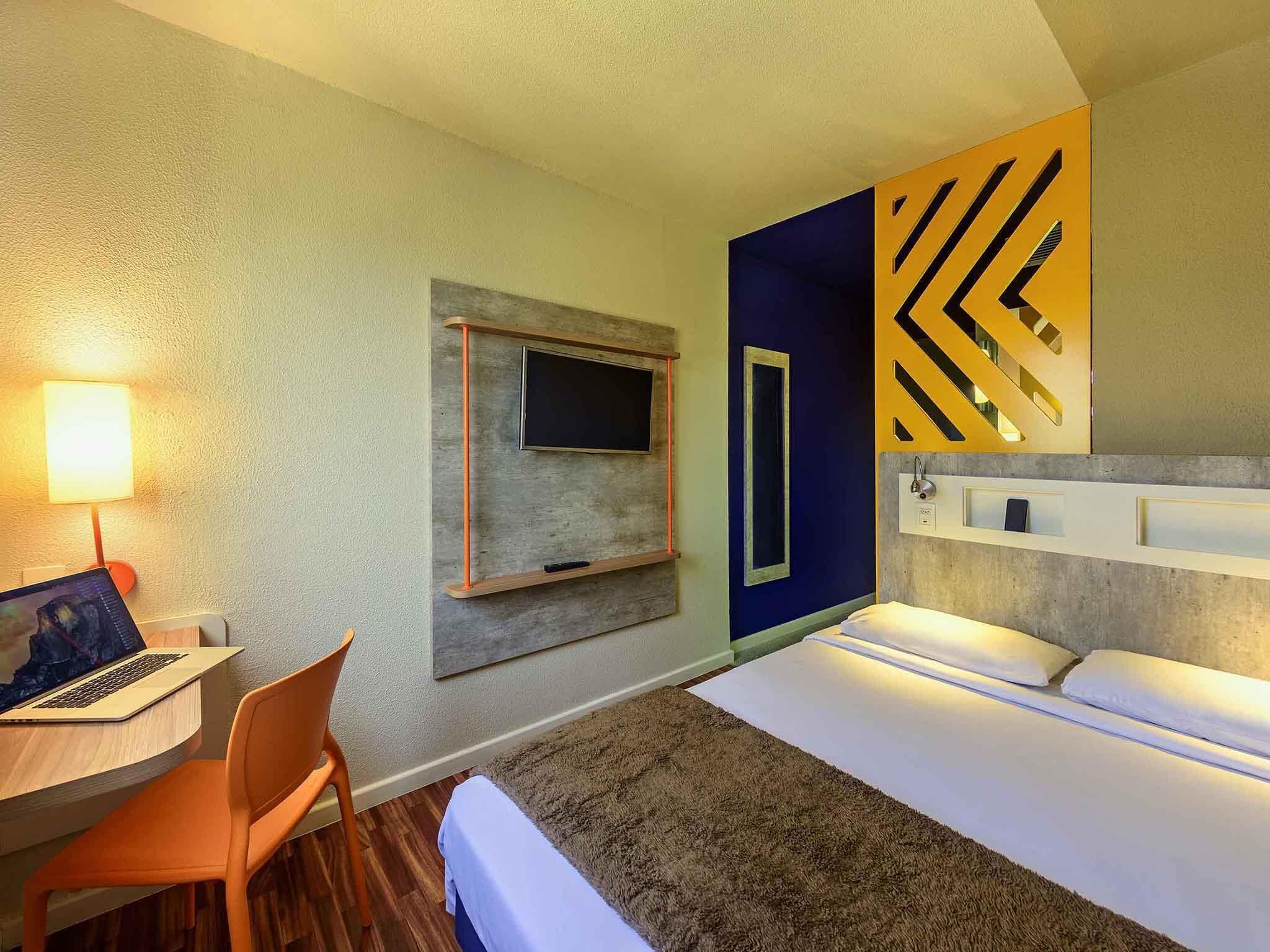 Quarto de hotel com uma cama, uma mesa com um laptop, uma cadeira, uma TV fixada na parede e um painel decorativo.