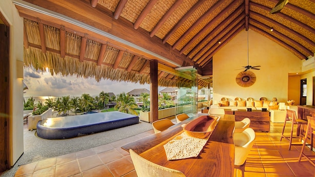 Uma sala de jantar ampla com uma grande mesa de madeira, assentos confortáveis e vista para uma piscina e palmeiras.