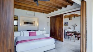 Quarto moderno com uma cama grande, vigas de madeira no teto e um espaço de jantar com cadeiras e mesa de madeira.