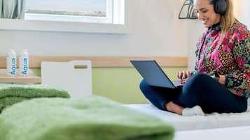 Uma pessoa sentada na cama com um laptop, uma janela com vista para a cidade e duas garrafas com a etiqueta 'Agua' em uma mesa de cabeceira.