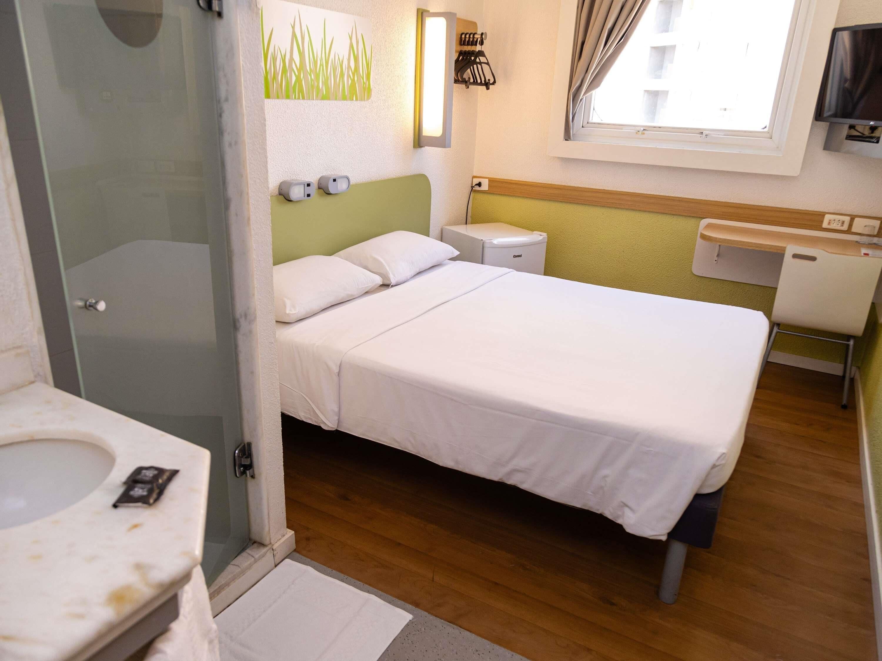 Quarto de hotel com cama, mesa de cabeceira, pequena escrivaninha, janela com cortinas e chuveiro.