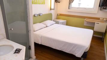 Quarto de hotel com cama, mesa de cabeceira, pequena escrivaninha, janela com cortinas e chuveiro.