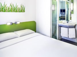 Quarto de hotel com cama, cabeceira verde, luminária na parede, banheiro com box de vidro e pia com toalhas.