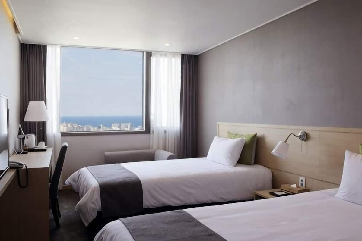 Quarto de hotel com duas camas, uma escrivaninha, uma cadeira, uma luminária e vista para a cidade.