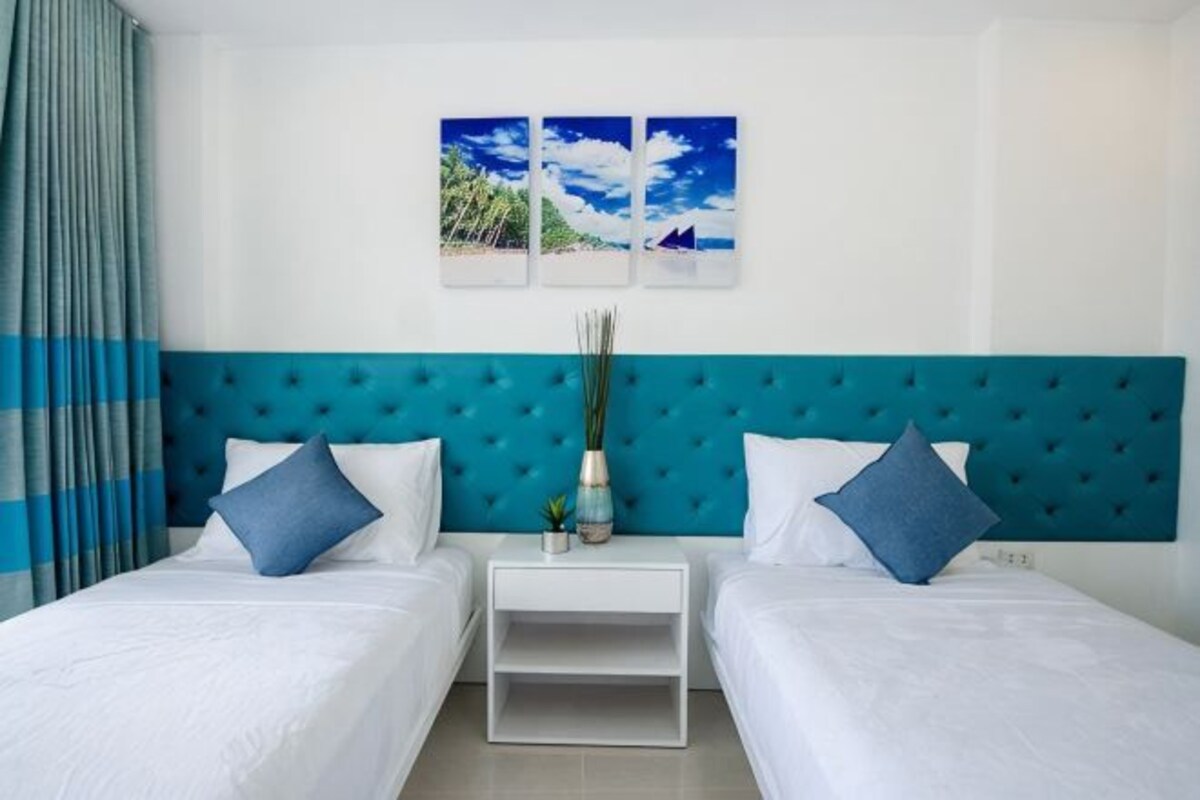 Quarto de hotel com duas camas, cabeceira verde-água, roupa de cama branca e duas fotografias emolduradas na parede.