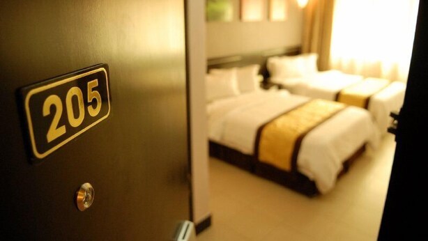 Quarto de hotel porta com a inscrição '205' e duas camas com roupa de cama branca e detalhes dourados.
