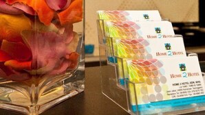 Uma área de recepção com um vaso de vidro transparente cheio de pétalas coloridas, um balcão de recepção e um monitor exibindo uma tela.