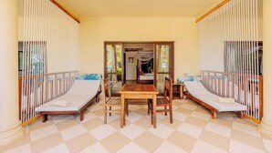 Um quarto com piso xadrez, uma mesa de madeira, duas cadeiras de madeira, um banco de madeira e uma porta de vidro que dá para outro cômodo.