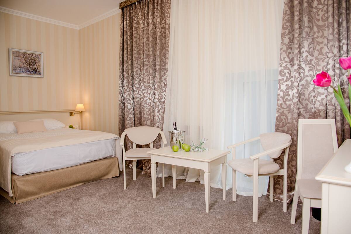 Quarto de hotel com cama, duas cadeiras, uma mesa pequena com um vaso de flores e um abajur.