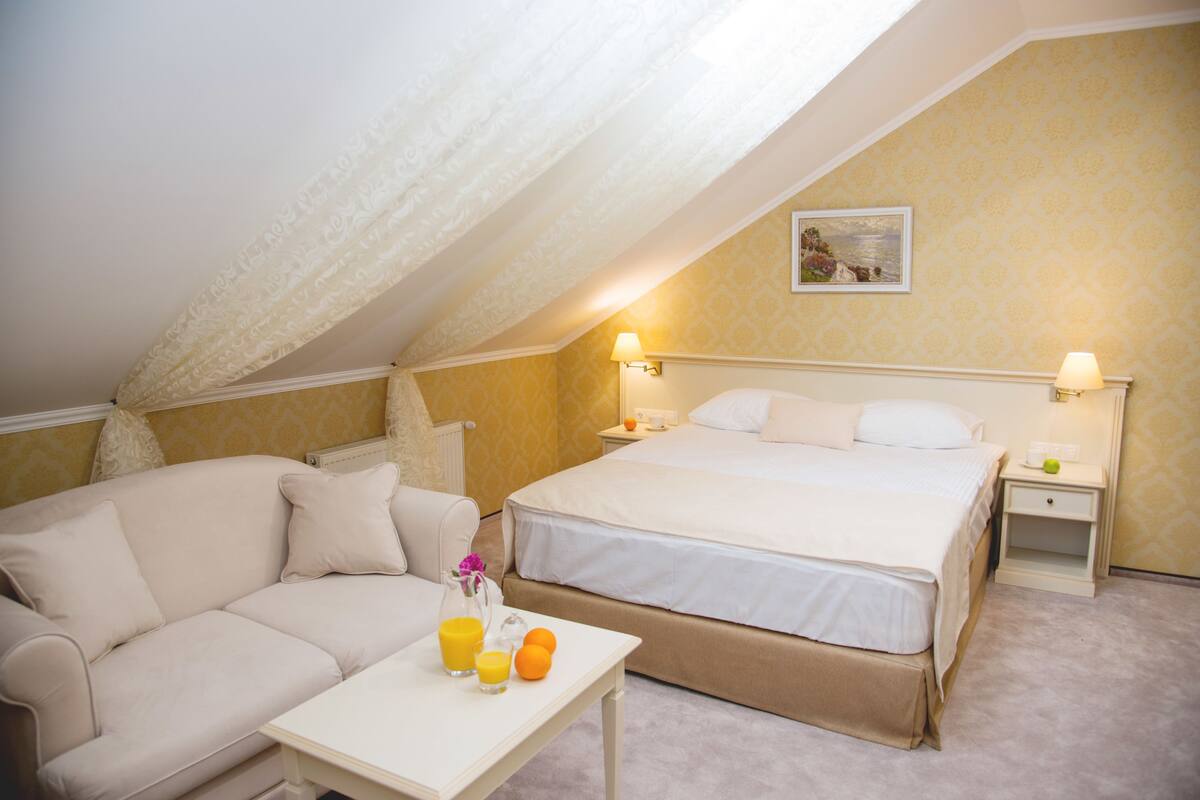 Quarto de hotel com uma cama, um sofá, uma mesa com um copo de suco de laranja e uma luminária na mesinha de cabeceira.