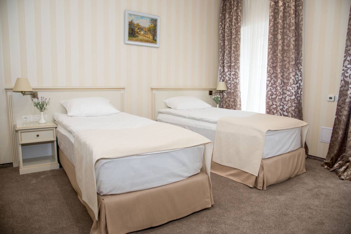Quarto de hotel com duas camas, mesa de cabeceira com abajur, pequena mesa com um vaso de flores e um quadro na parede.