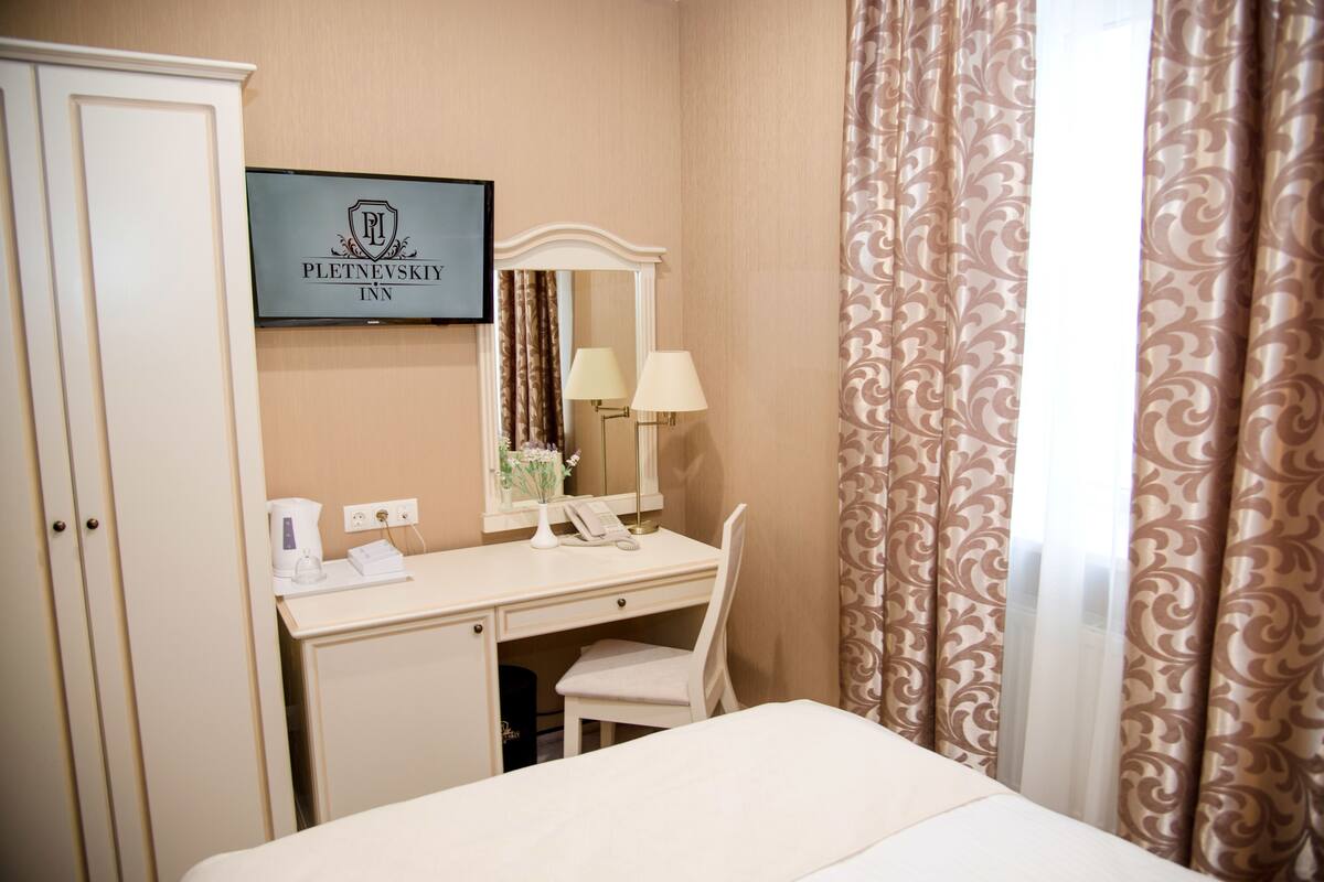 Quarto de hotel com cama, escrivaninha com cadeira, espelho, televisão e janela com cortinas.