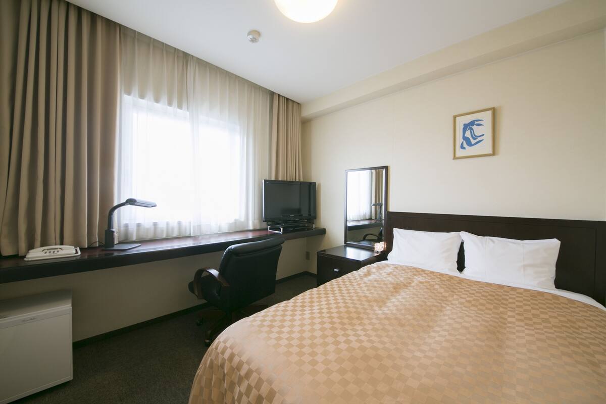 Quarto de hotel com cama, escrivaninha com computador, cadeira, televisão e janela com cortinas.