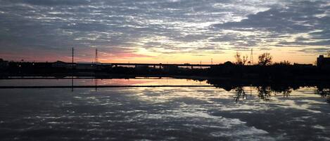 Um pôr do sol sobre a água, com uma ponte e estruturas industriais ao fundo.