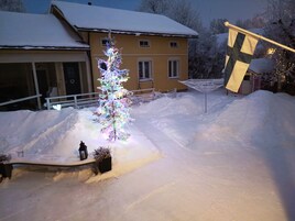 Paisagem coberta de neve com um pinheiro de Natal iluminado e uma casa ao fundo.