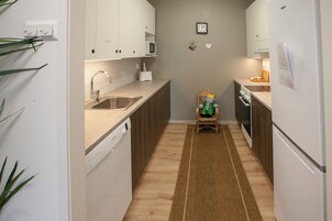 Uma cozinha compacta com armários brancos, micro-ondas, pia e lava-louças. Há um tapete pequeno no chão e um relógio de parede decorativo.