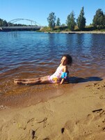 Criança sentada em uma praia arenosa perto da água, com uma ponte ao fundo.
