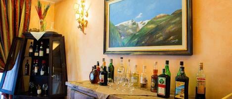 Uma área de bar com balcão de madeira, garrafas de bebidas e copos de vinho. Uma pintura emoldurada de uma paisagem montanhosa está pendurada acima do balcão.