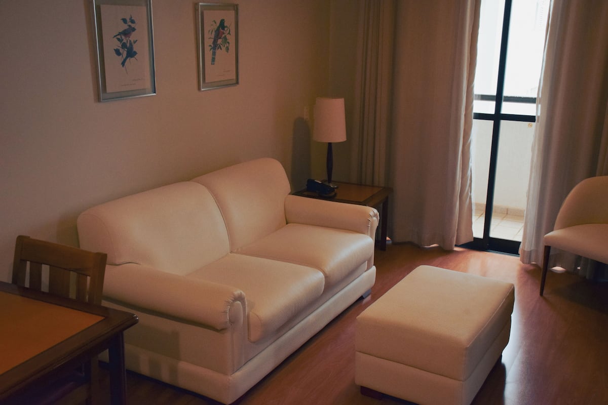 Sala de estar com sofá, uma mesinha, abajur e uma cadeira. Duas quadros emoldurados na parede.