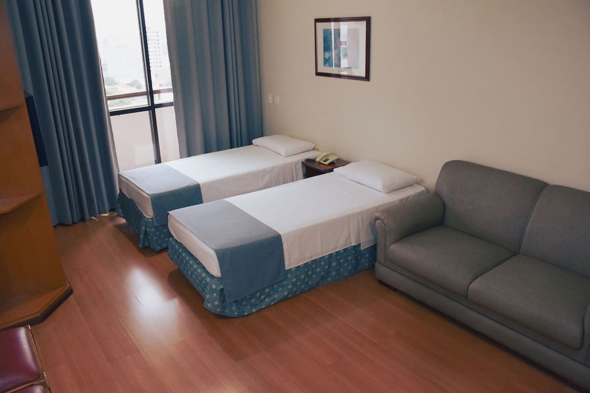 Quarto de hotel com duas camas de solteiro, um sofá cinzento, piso de madeira e uma janela com cortinas.