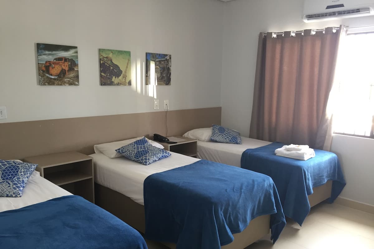 Quarto de hotel com duas camas, cada uma com colcha azul e almofadas combinando, uma mesa de cabeceira com telefone e uma janela com cortinas.