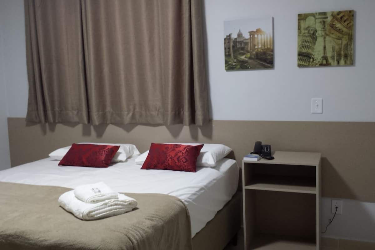 Quarto de hotel com uma cama, duas almofadas vermelhas, um cobertor branco, uma cabeceira marrom, uma mesa de cabeceira com um telefone e duas obras de arte nas paredes retratando edifícios históricos.