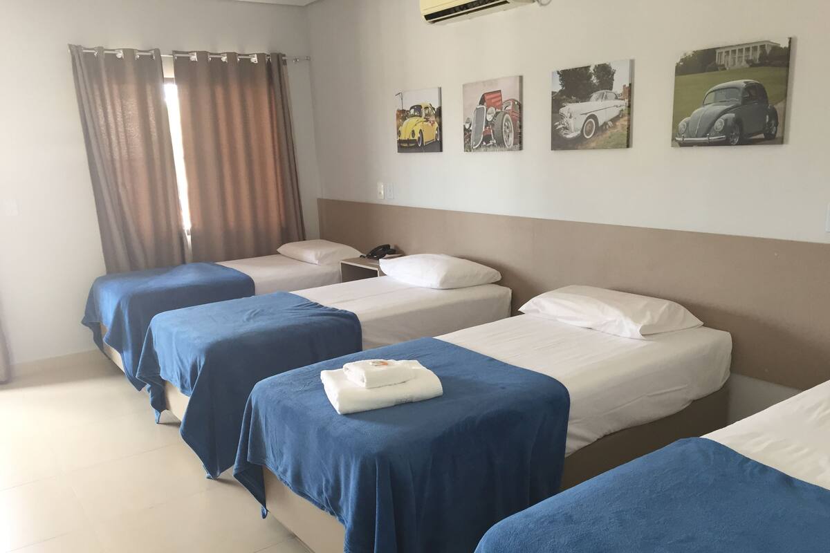 Quarto de hotel com quatro camas de solteiro, cada uma com colcha azul e travesseiros brancos. Há uma janela com cortinas, ar-condicionado fixado na parede e três quadros com imagens de carros antigos.