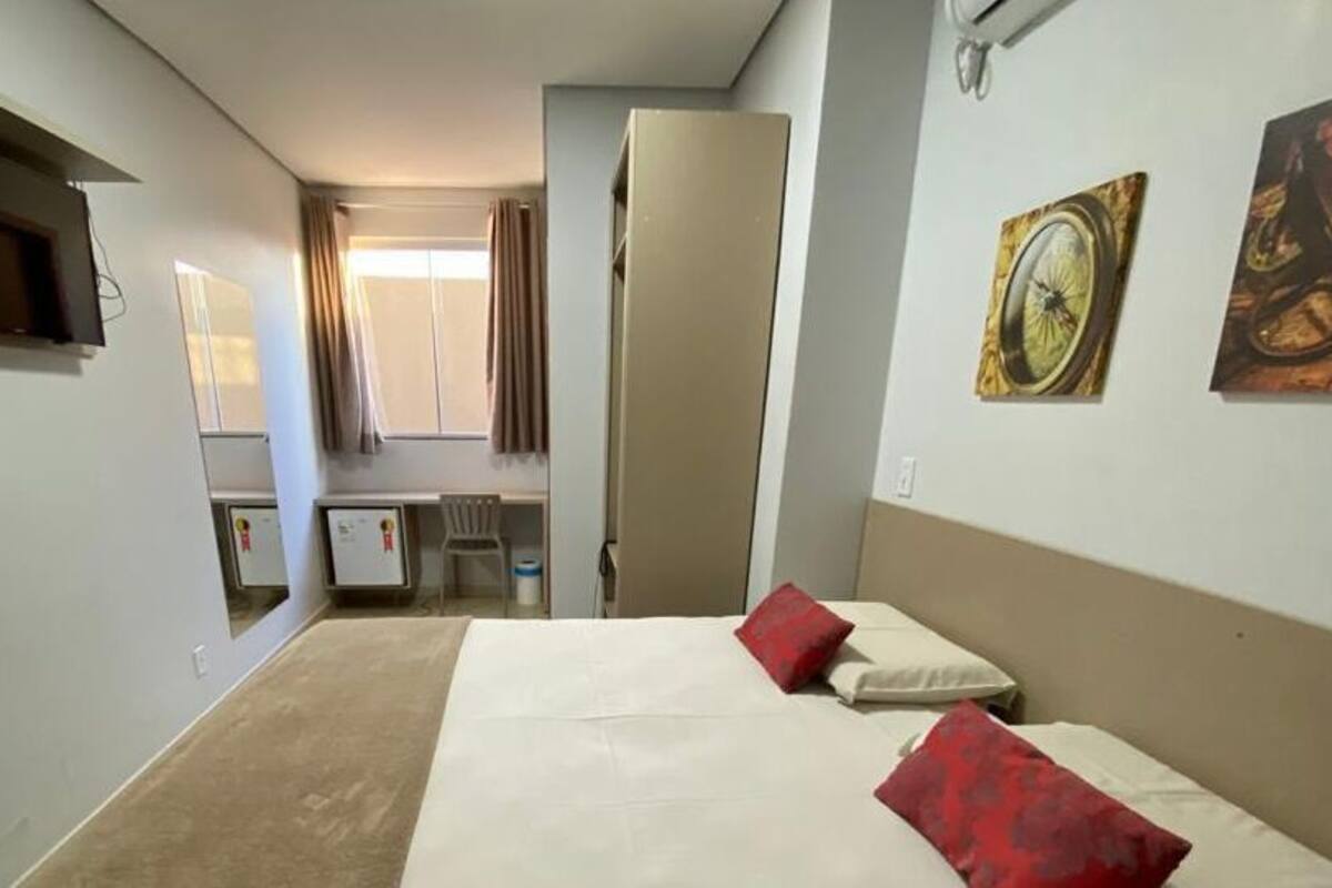 Quarto de hotel com cama, escrivaninha, cadeira e janela com cortinas.