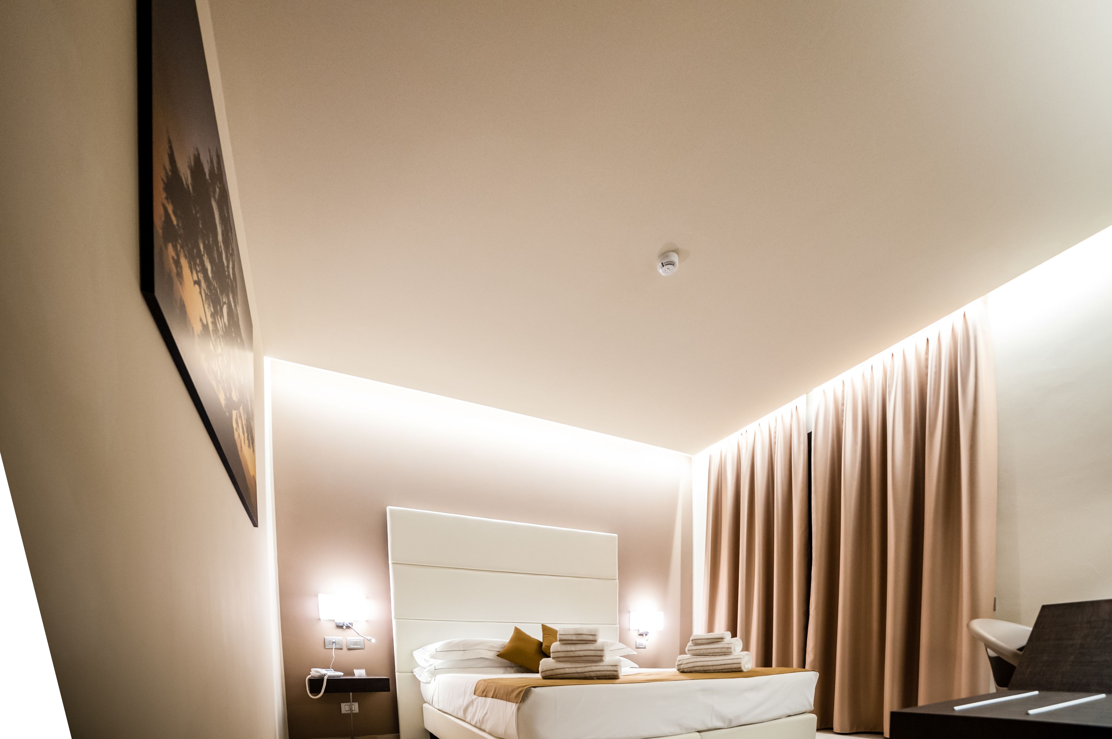 Quarto de hotel moderno com uma cama bem arrumada, uma mesa de cabeceira com abajur e uma janela grande com cortinas.