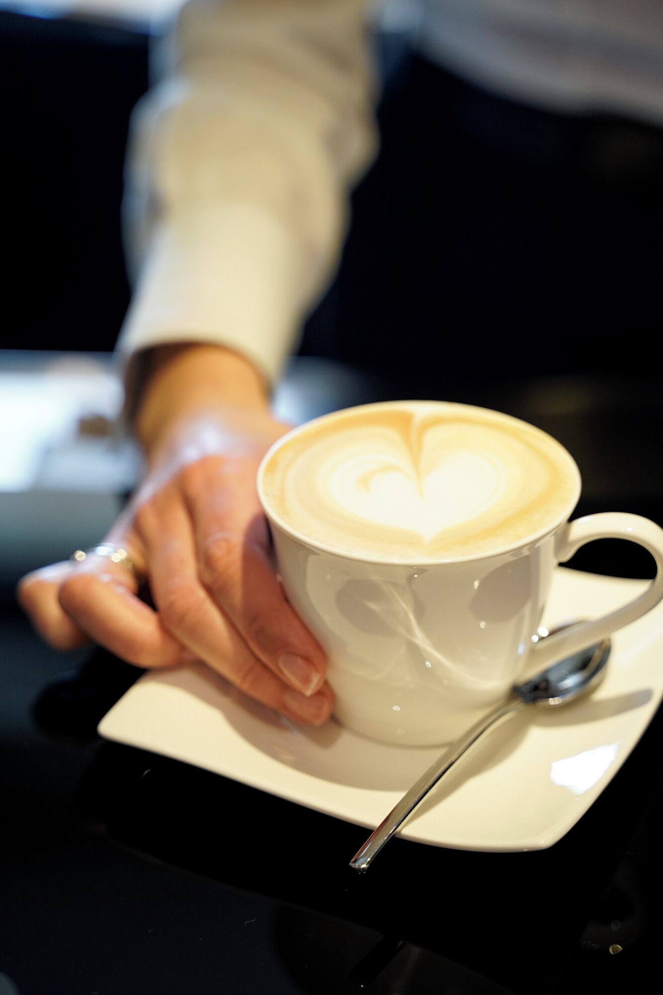 Uma chávena de café com arte de latte num pires, uma colher e a mão de alguém a segurar a chávena.