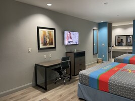 Quarto de hotel com escrivaninha, cadeira, duas camas, televisão e espelho no banheiro.